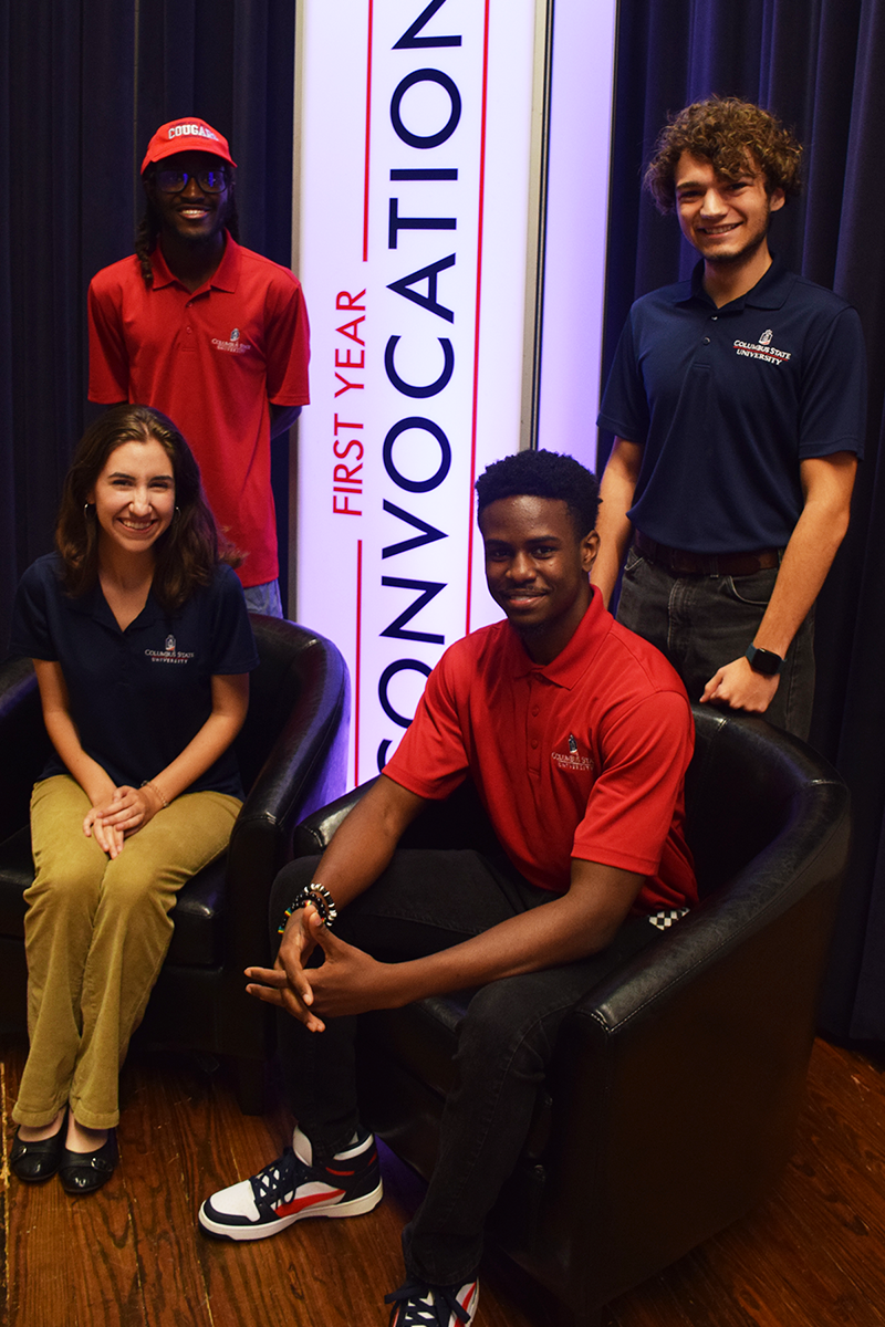 group of 2 students seated and 2 students standing in front of a pillar that reads First Year ...